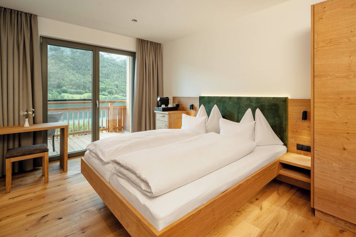 Quarto de hotel moderno com uma cama grande, uma escrivaninha e vista para uma área arborizada.