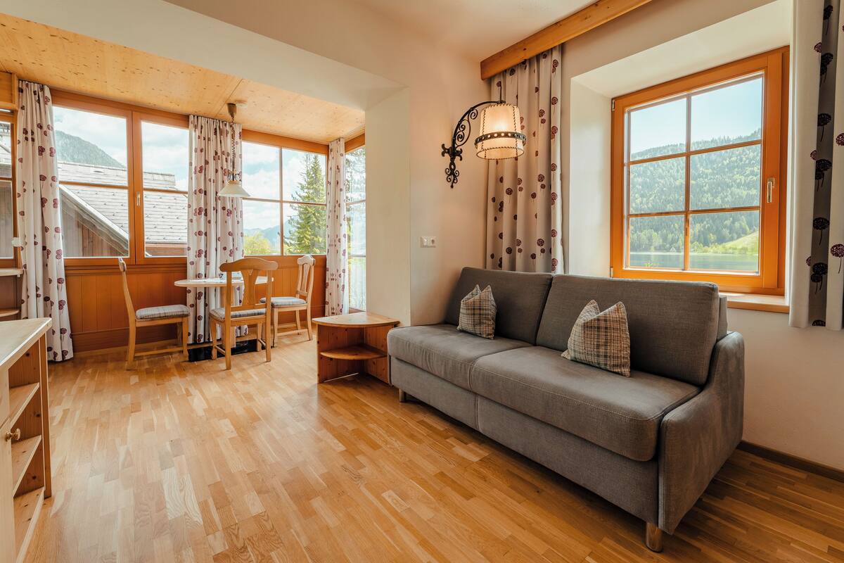 Um quarto aconchegante com sofá, piso de madeira, uma mesa com cadeiras, janela com cortinas e uma luminária na parede.