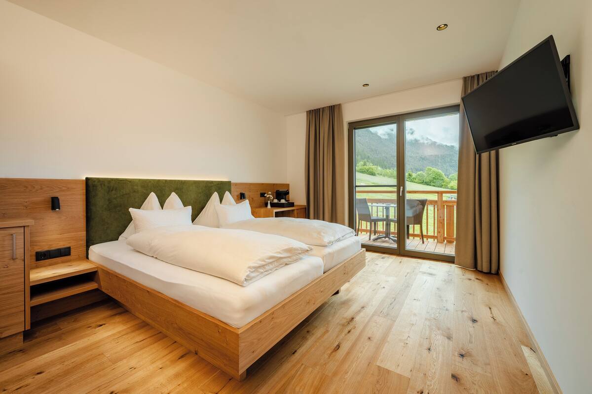 Quarto de hotel moderno com cama de madeira, televisão na parede e varanda com vista para as montanhas.
