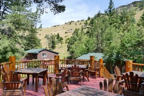 Um deck de madeira com mesas e cadeiras, com vista para uma encosta arborizada com cabanas.