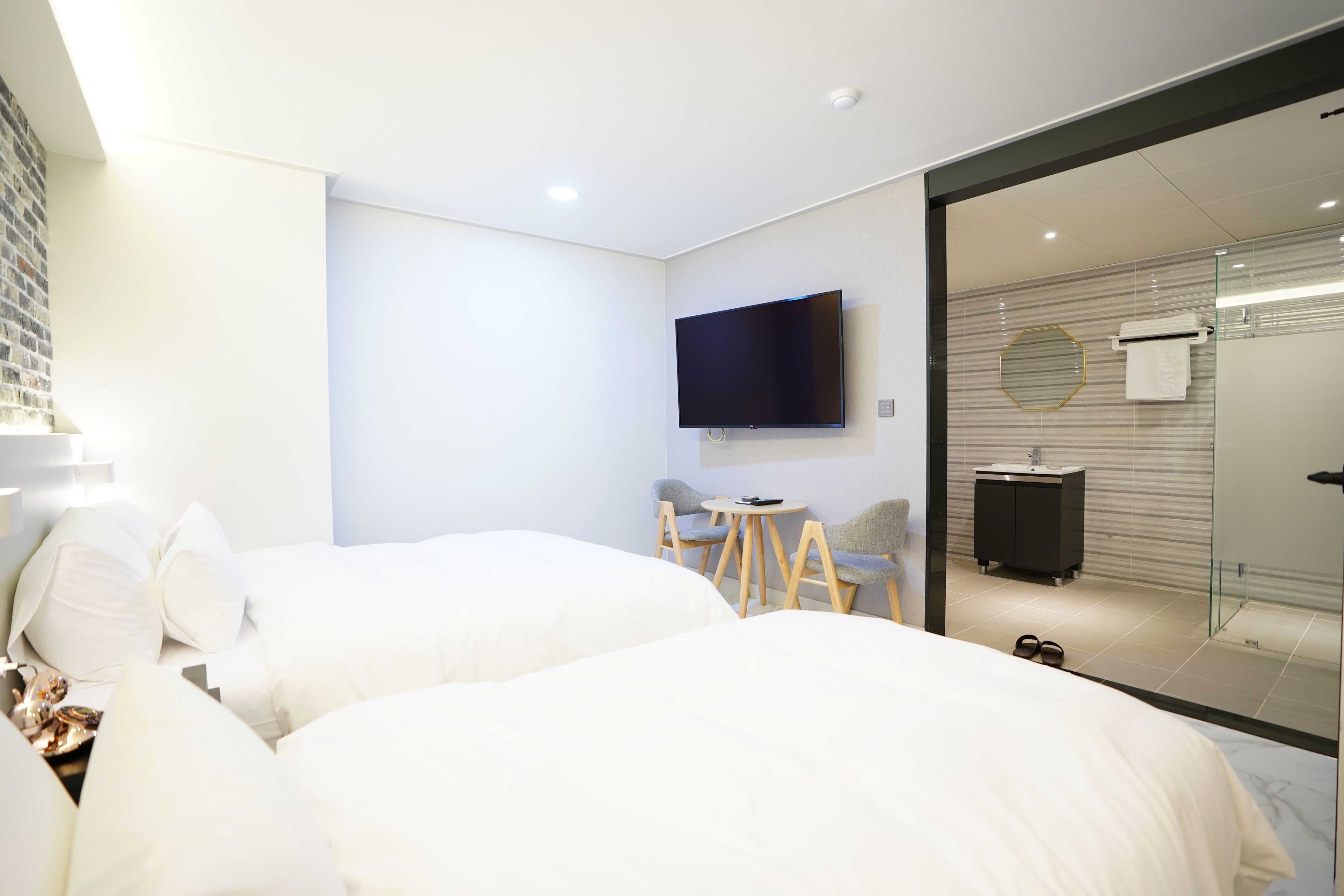 Quarto de hotel moderno com duas camas, televisão de tela plana, uma pequena mesa com cadeiras e um painel de vidro separando o quarto do banheiro.