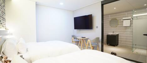 Quarto de hotel moderno com duas camas, televisão de tela plana, uma pequena mesa com cadeiras e um painel de vidro separando o quarto do banheiro.