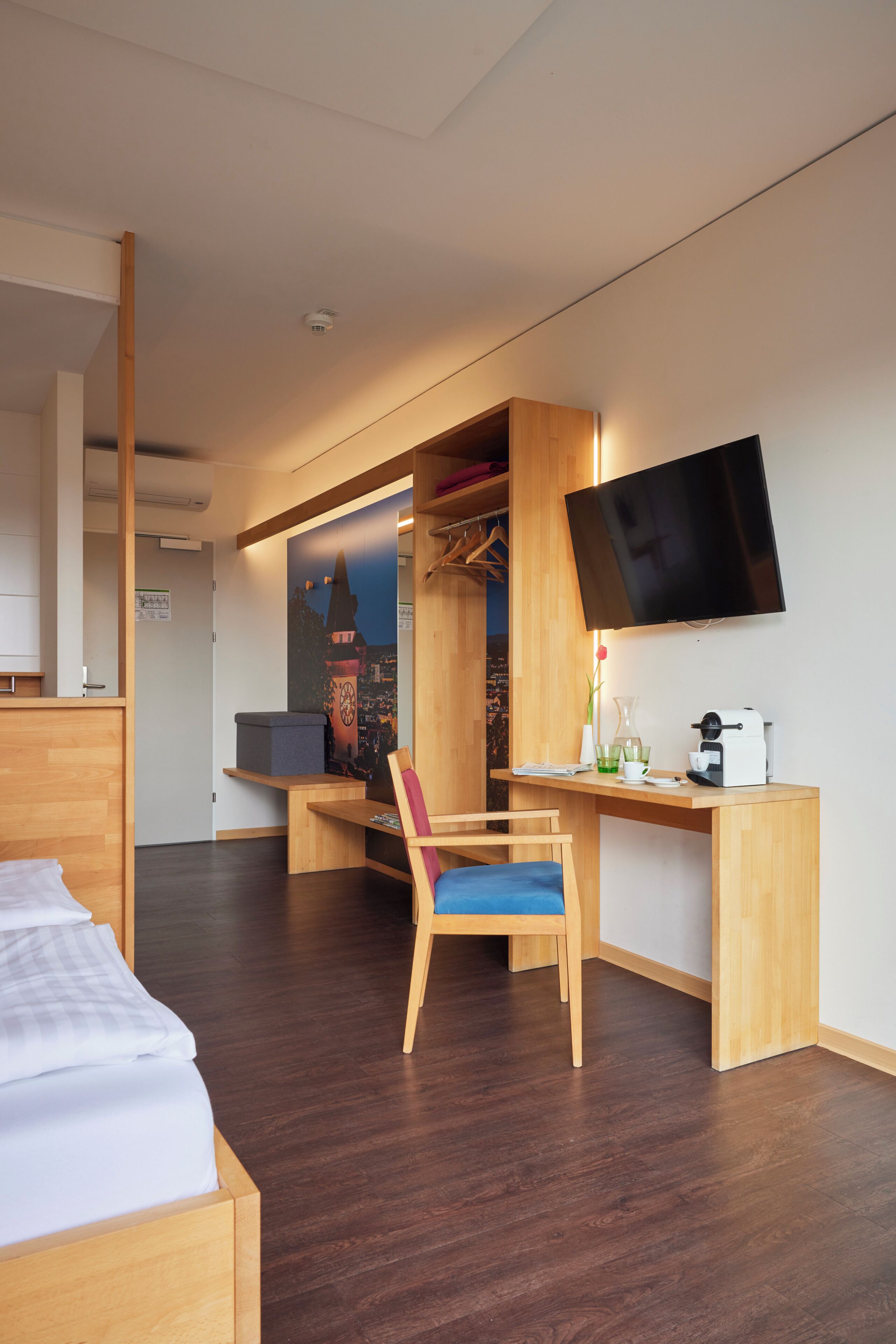 Quarto de hotel pequeno com uma cama, uma escrivaninha com cadeira, televisão e um cartaz de uma paisagem urbana.