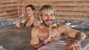 Uma cabana de madeira com hidromassagem, uma mulher e um homem segurando copos de vinho, e uma placa indicando que não é permitido fumar.
