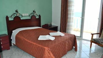 Cama bem arrumada com colcha estampada e duas toalhas, mesa de cabeceira de madeira com um copo, e uma cadeira com almofada estampada.