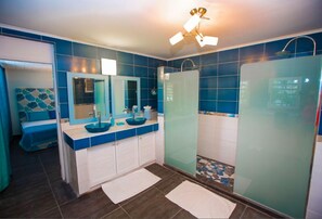 Casa de banho com parede em azulejos azuis, cabine de duche em vidro, lavatório branco com cuba azul e ventoinha no teto.