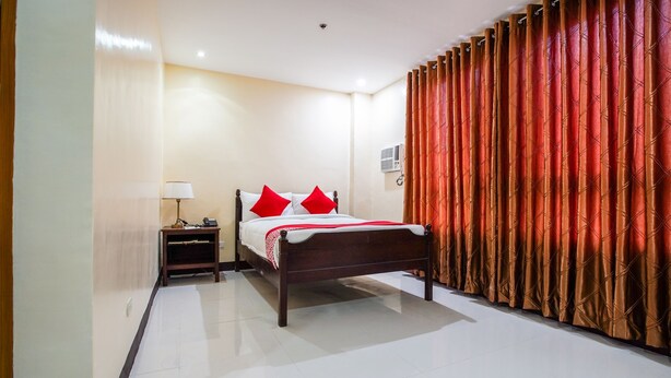 Quarto de hotel com cama, mesa de cabeceira, abajur e cortina.
