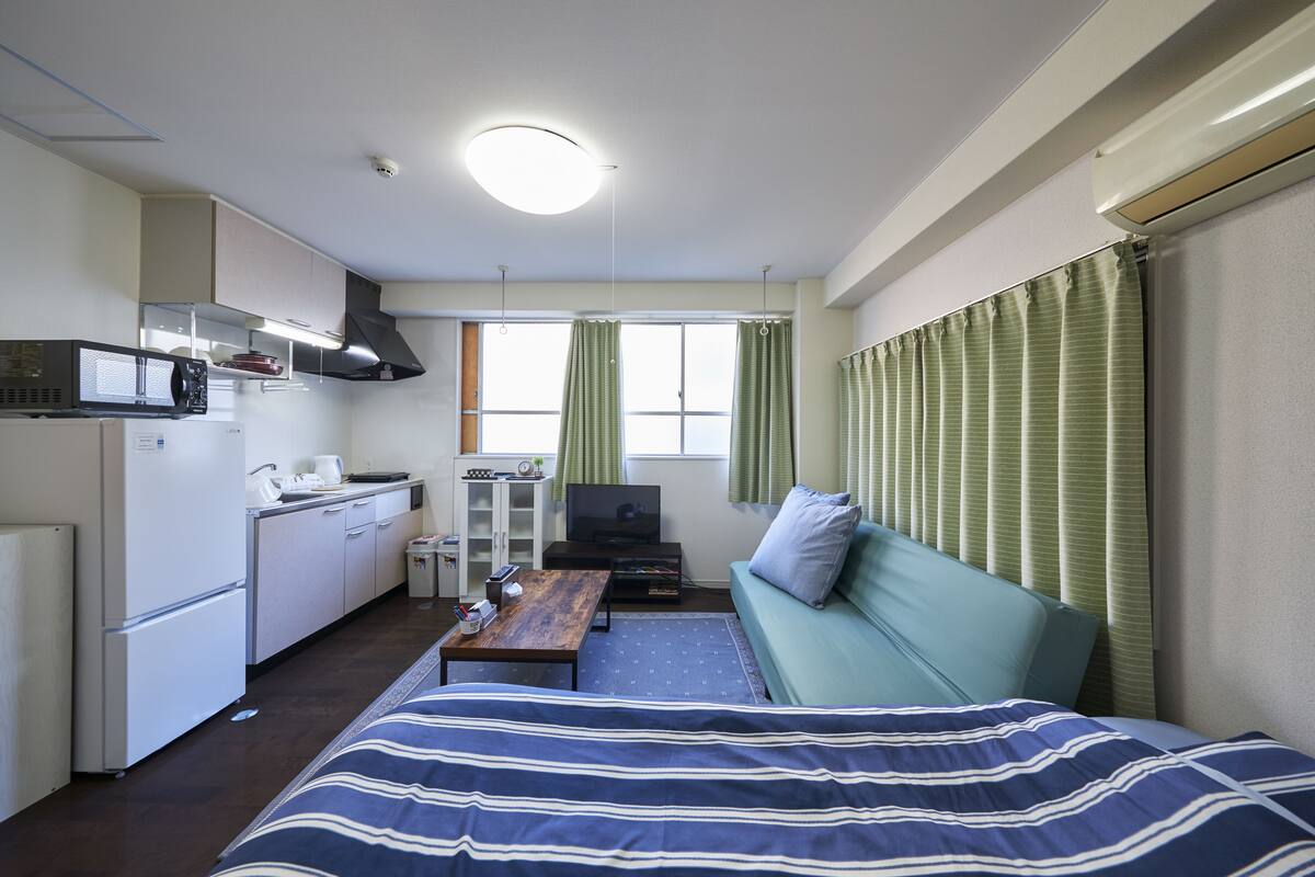 Quarto de hotel pequeno com cozinha compacta, sofá, televisão e janela com cortinas.