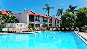 Uma piscina com espreguiçadeiras e um edifício branco com telhados vermelhos ao fundo.