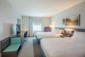 Quarto de hotel com duas camas, uma escrivaninha, uma cadeira, uma televisão, um quadro e uma janela com cortinas.