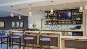 Uma área de bar moderna com um balcão comprido, vários bancos altos e uma variedade de garrafas e copos nas prateleiras.