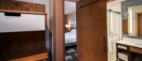 Quarto de hotel moderno com porta deslizante para o banheiro, cama com roupa de cama branca e um espelho grande sobre a pia.