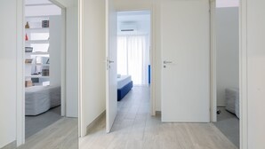 Corredor que dá para um quarto com cama, mesa de cabeceira e janela com cortinas.