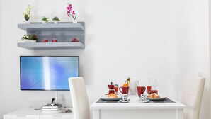 Uma sala de estar moderna com televisão de tela plana, mobiliário branco e um espaço de jantar com itens de café da manhã.