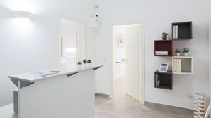 Uma área de recepção moderna e bem iluminada, com uma mesa branca, uma pequena planta e uma prateleira na parede com livros e um porta-retrato.