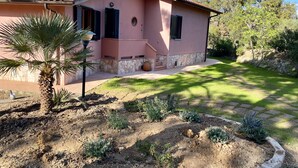 Uma villa rosa com base de pedra, uma palmeira e um jardim com diversas plantas.