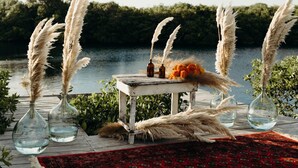Ambiente rústico ao ar livre com mesa de madeira, vasos de capim-dos-pampas, tapete estampado vermelho e um lago e vegetação ao fundo.