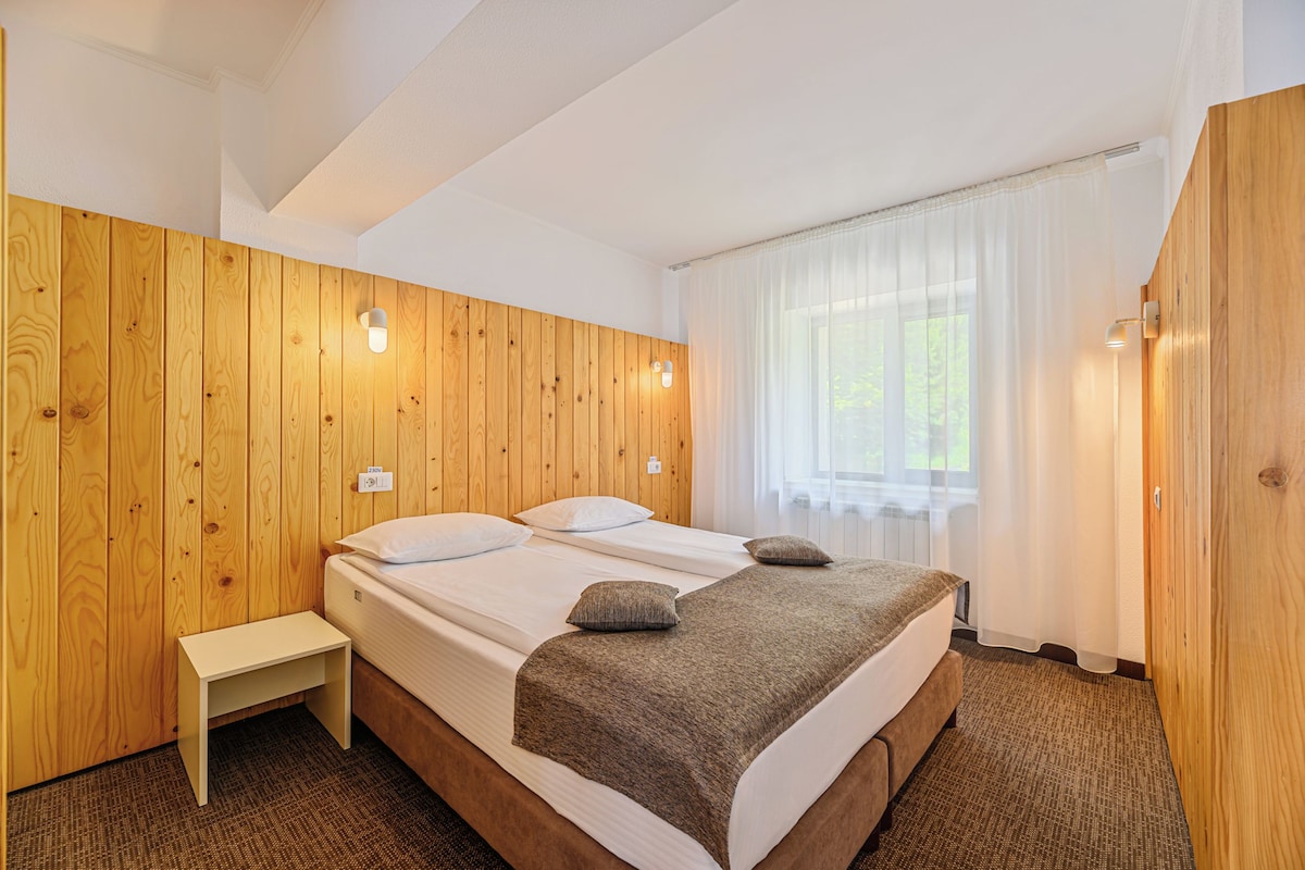 Quarto de hotel com parede de madeira, cama com roupa de cama branca, mesa de cabeceira e janela com cortinas.