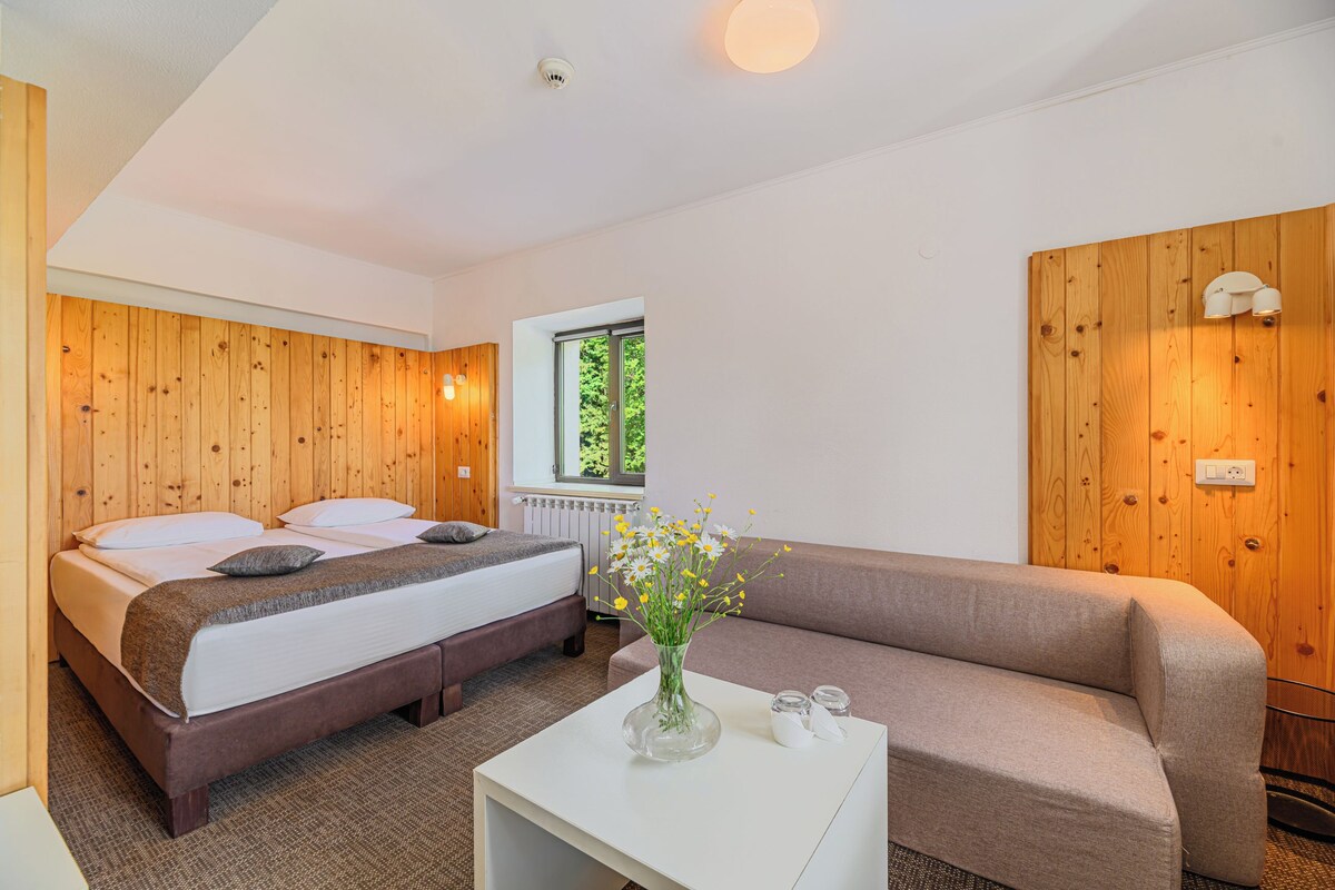 Quarto de hotel com cama, sofá, mesa pequena e janela vista para o verde.