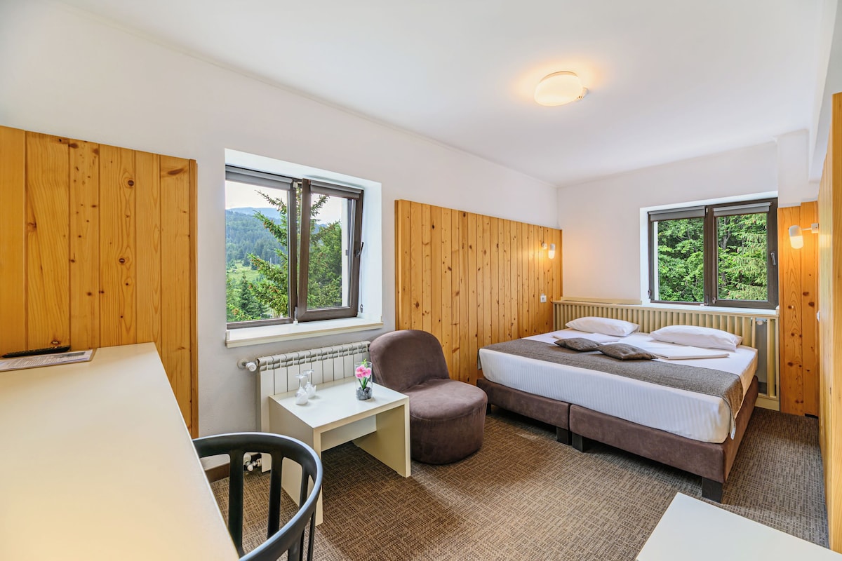 Quarto com cama, escrivaninha, cadeira e janela com vista para área verde.