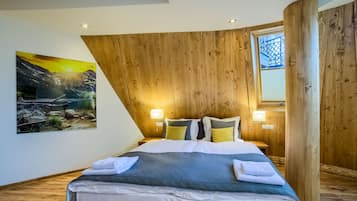 Um quarto com teto de madeira, cama com roupa de cama branca e cinza, duas luminárias de mesa e uma pintura de paisagem montanhosa na parede.