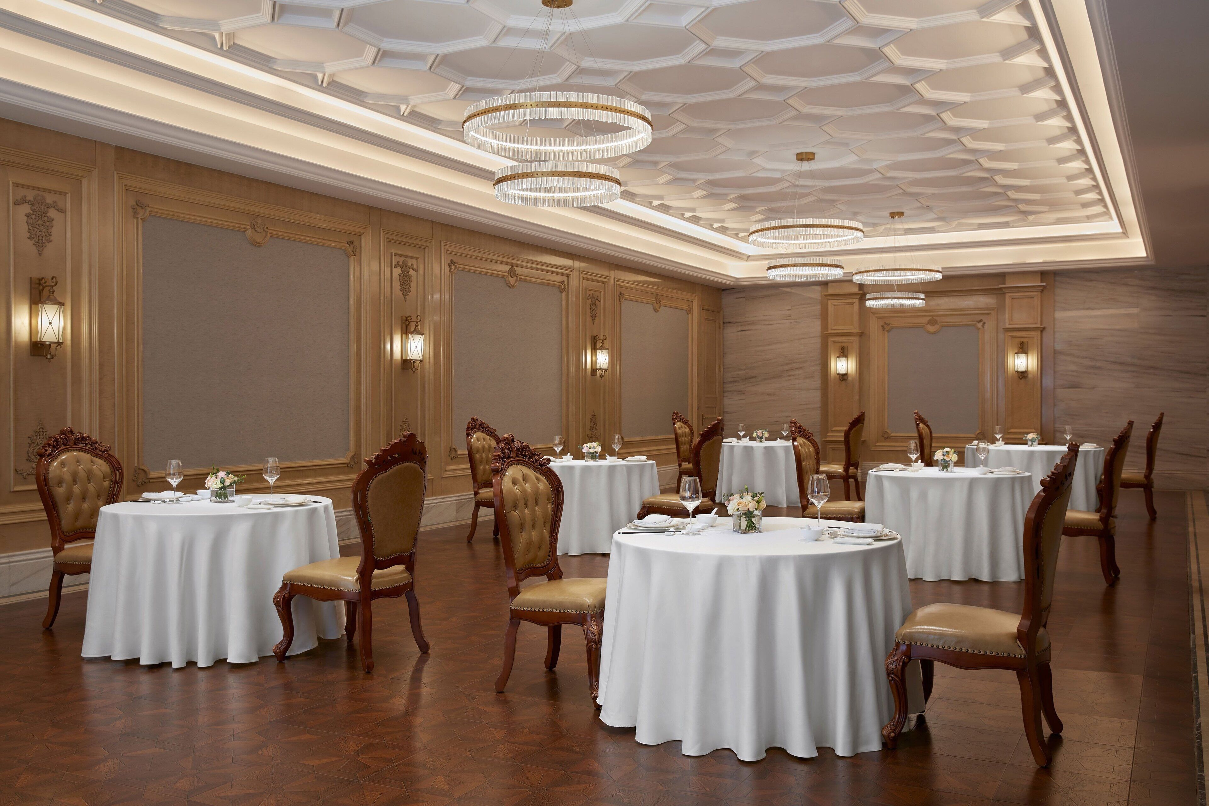 Uma sala de jantar formal com mesas redondas, cadeiras ricamente decoradas e lustres.