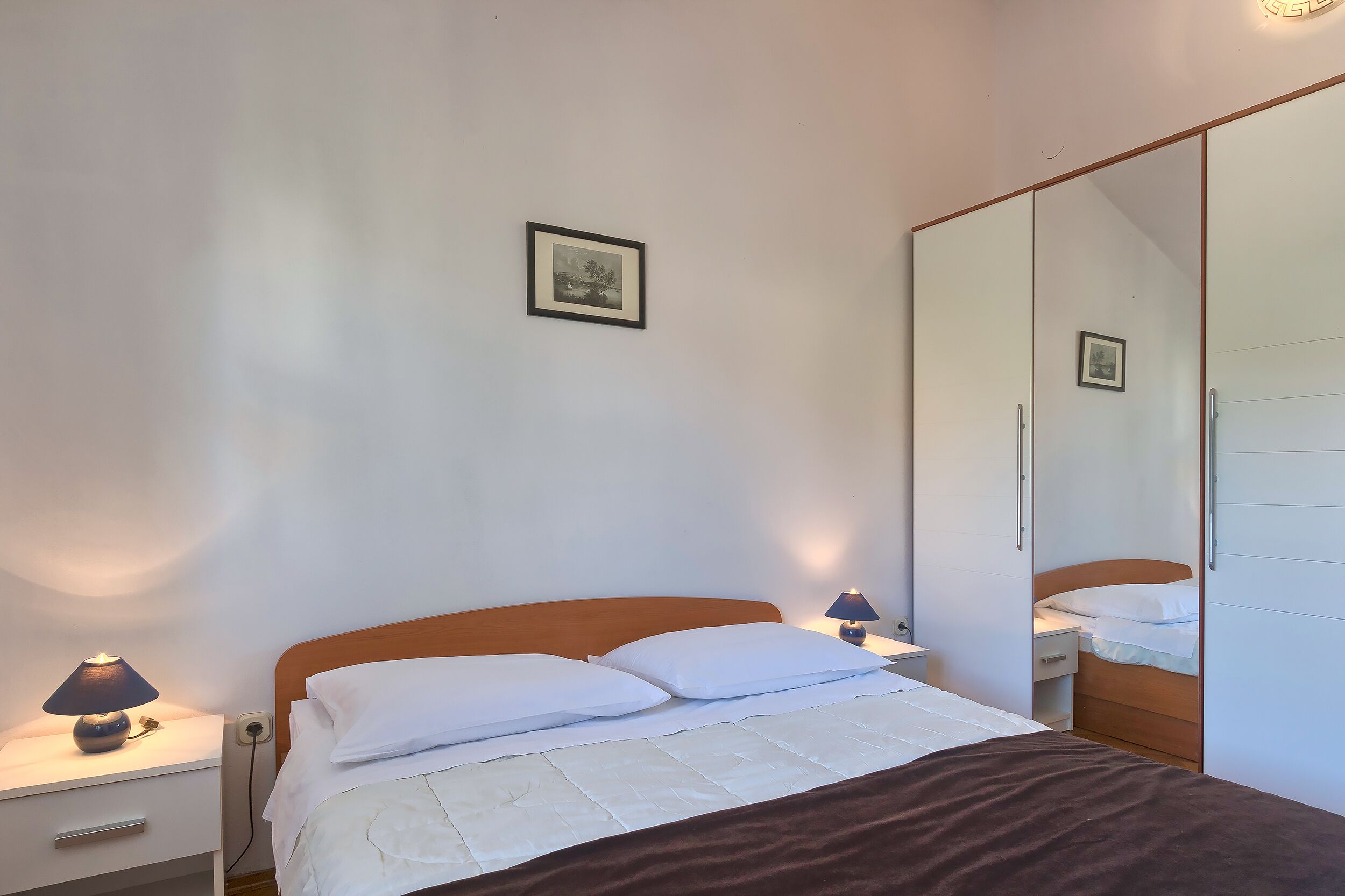 Uma cama bem arrumada com roupa de cama branca e um cobertor marrom, uma mesa de cabeceira com abajur, uma pequena mesa de cabeceira com abajur e um guarda-roupa com espelho.
