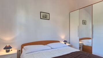 Uma cama bem arrumada com roupa de cama branca e um cobertor marrom, uma mesa de cabeceira com abajur, uma pequena mesa de cabeceira com abajur e um guarda-roupa com espelho.
