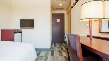 Quarto de hotel com cama, escrivaninha, cadeira, luminária, televisão e porta que dá para outro espaço.