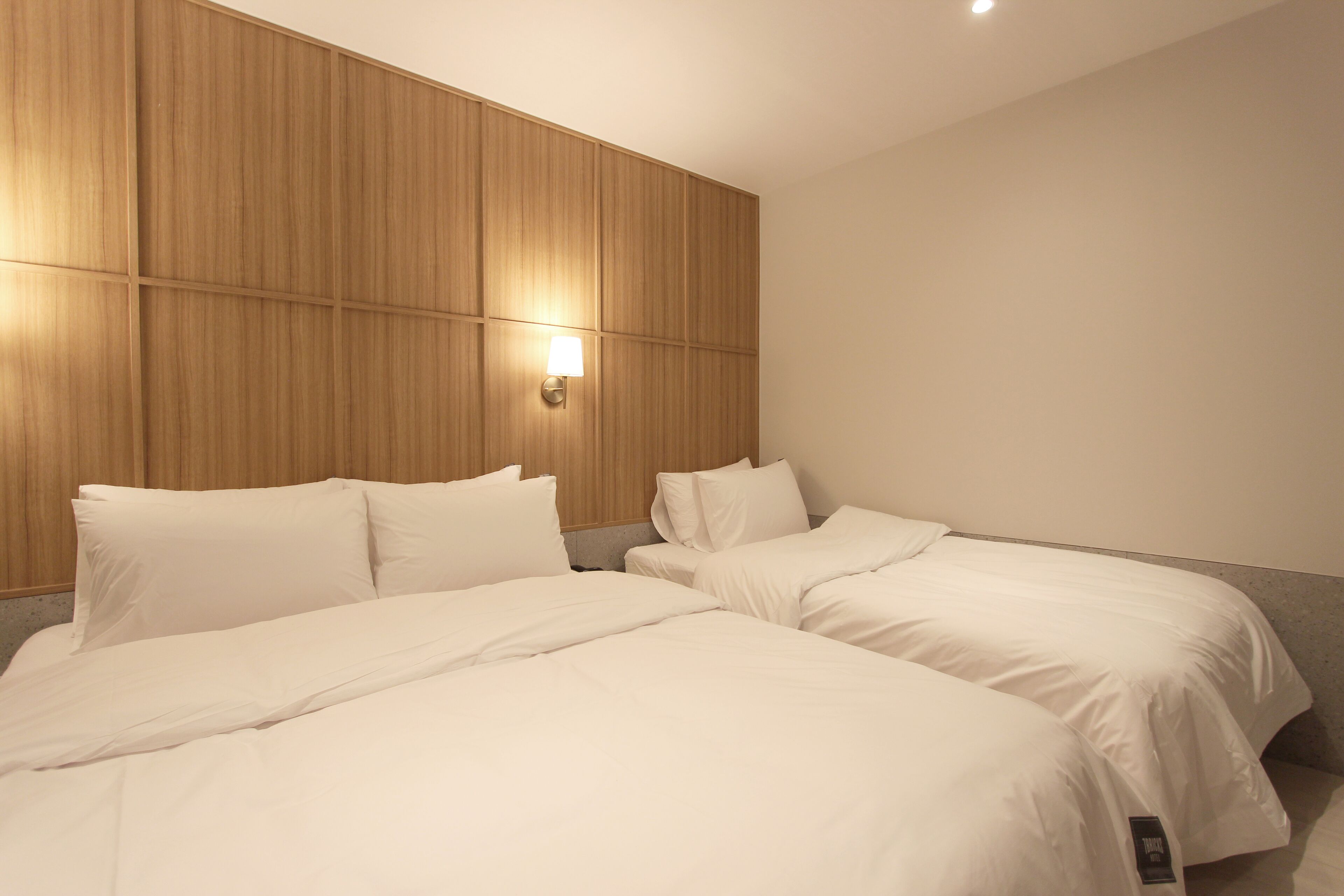 Quarto de hotel com duas camas, painéis de madeira na parede e luminária fixada na parede.