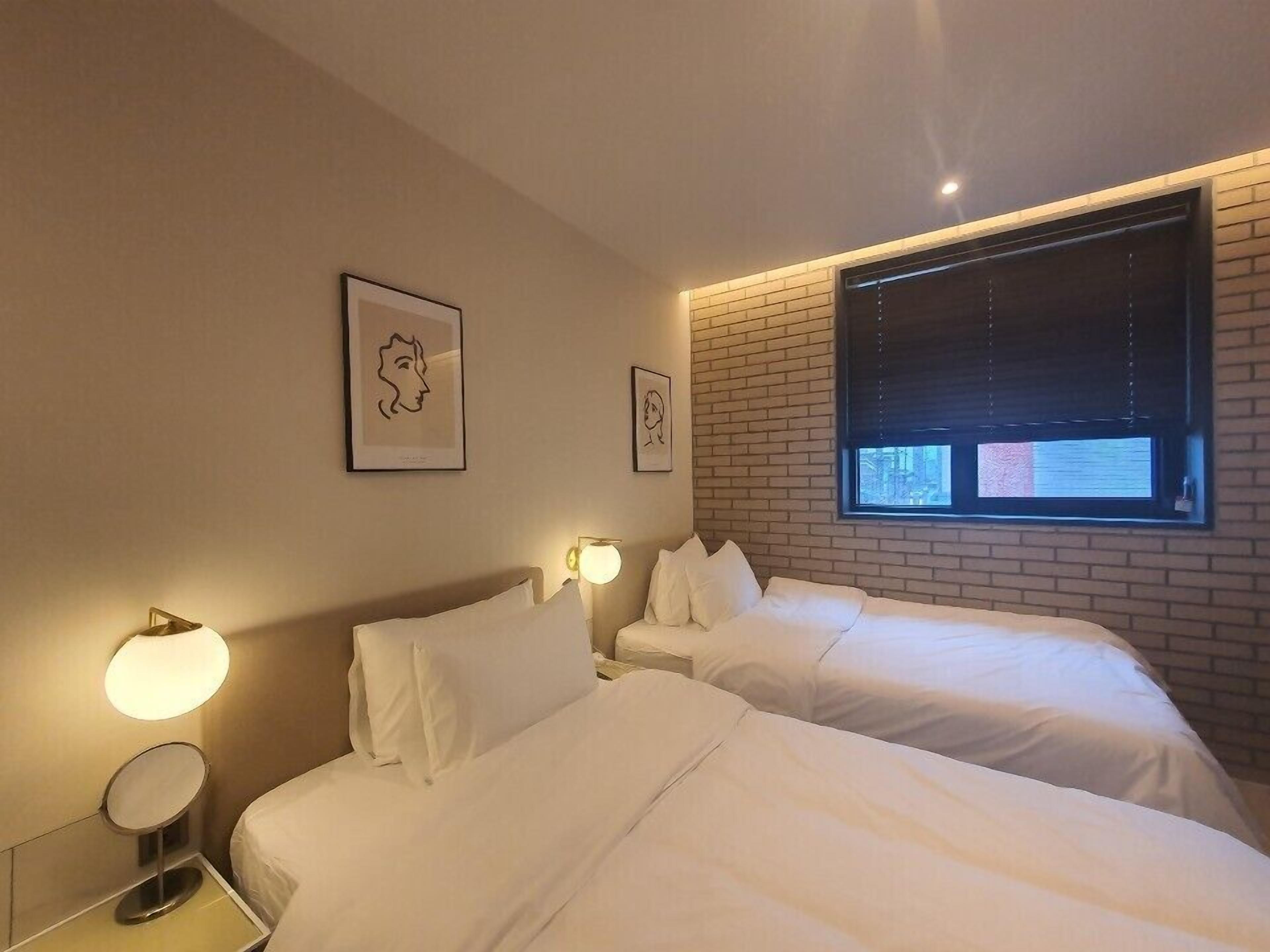 Quarto de hotel com duas camas, parede de tijolos e janela com persianas.
