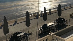 Vista da praia com área de refeições, incluindo mesas e guarda-sóis, com vista para o mar calmo.
