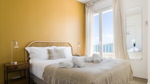 Cama bem arrumada com lençóis brancos e manta bege, luminária de cabeceira e mesa de cabeceira com gaveta.