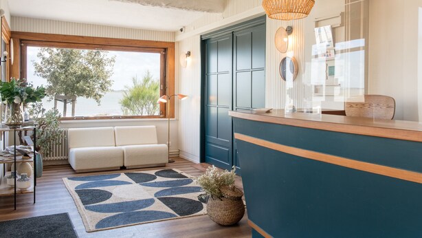Um quarto acolhedor com uma janela ampla, um banco de madeira, uma porta azul e um balcão de recepção.