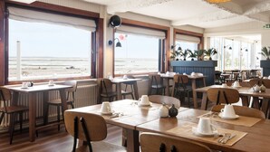 Um restaurante com mesas e cadeiras de madeira, grandes janelas com vista para a praia e plantas em vasos no parapeito.