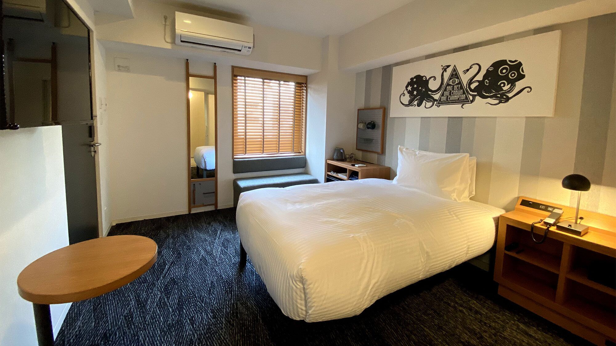 Quarto de hotel com cama, escrivaninha, cadeira, mesinha e janela com persianas.