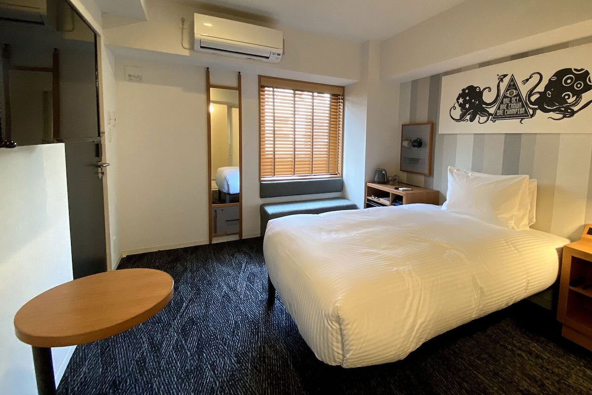 Quarto de hotel com cama, escrivaninha, cadeira, mesinha e janela com persianas.