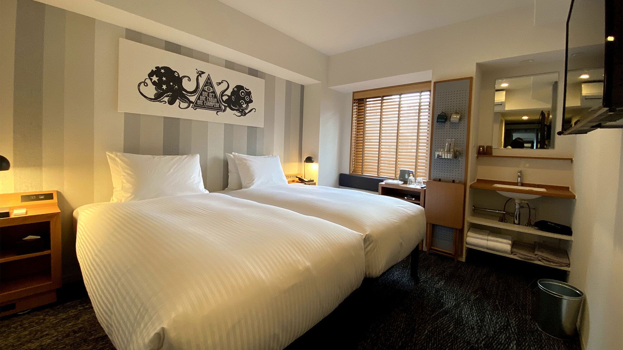 Quarto de hotel com duas camas, uma mesa, um espelho e uma janela com persianas.