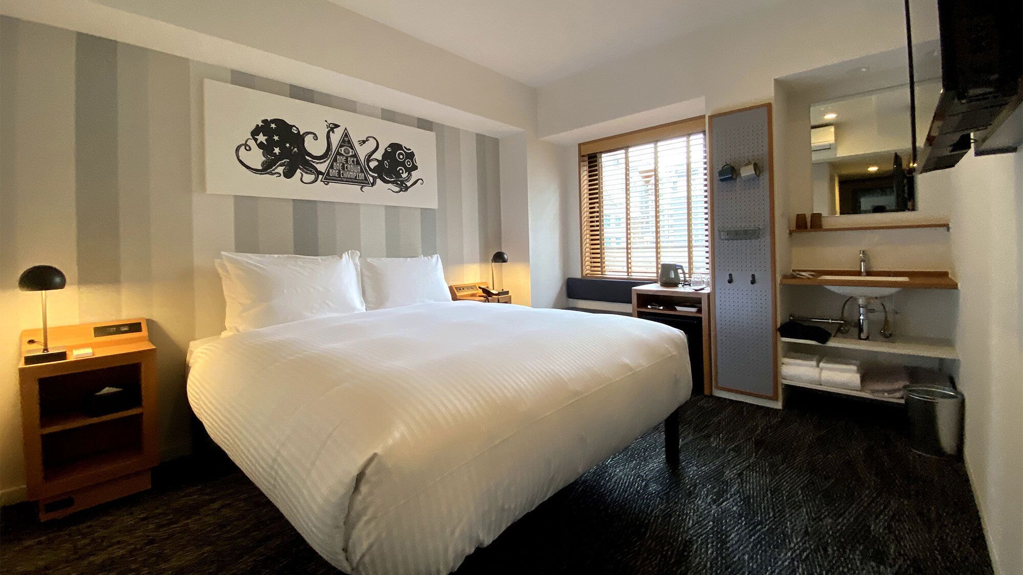 Quarto de hotel com uma cama grande, uma escrivaninha com computador, um espelho e um guarda-roupa.