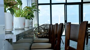 Uma área de refeições com vista para o mar, apresentando um balcão com plantas em vasos, uma cadeira estampada e cadeiras de madeira.
