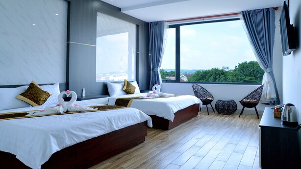 Quarto de hotel moderno com duas camas, uma janela grande com cortinas, uma televisão de tela plana e uma pequena mesa com um chaleira.