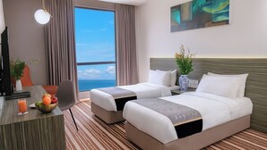 Quarto de hotel com duas camas, uma escrivaninha com uma cesta de frutas, um vaso com flores e uma janela grande com vista para o oceano.