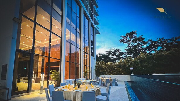 Uma área de refeições ao ar livre com mesas e cadeiras, piscina e um edifício com grandes janelas.