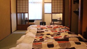 Um quarto tradicional japonês com tatame, futon com colcha estampada, portas deslizantes e uma pequena mesa com cadeira.