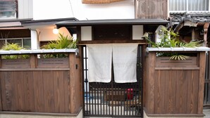 Um portão tradicional japonês com paredes de madeira e estrutura preta, que dá acesso a um edifício com telhado em telhas e uma pequena área de jardim com plantas.