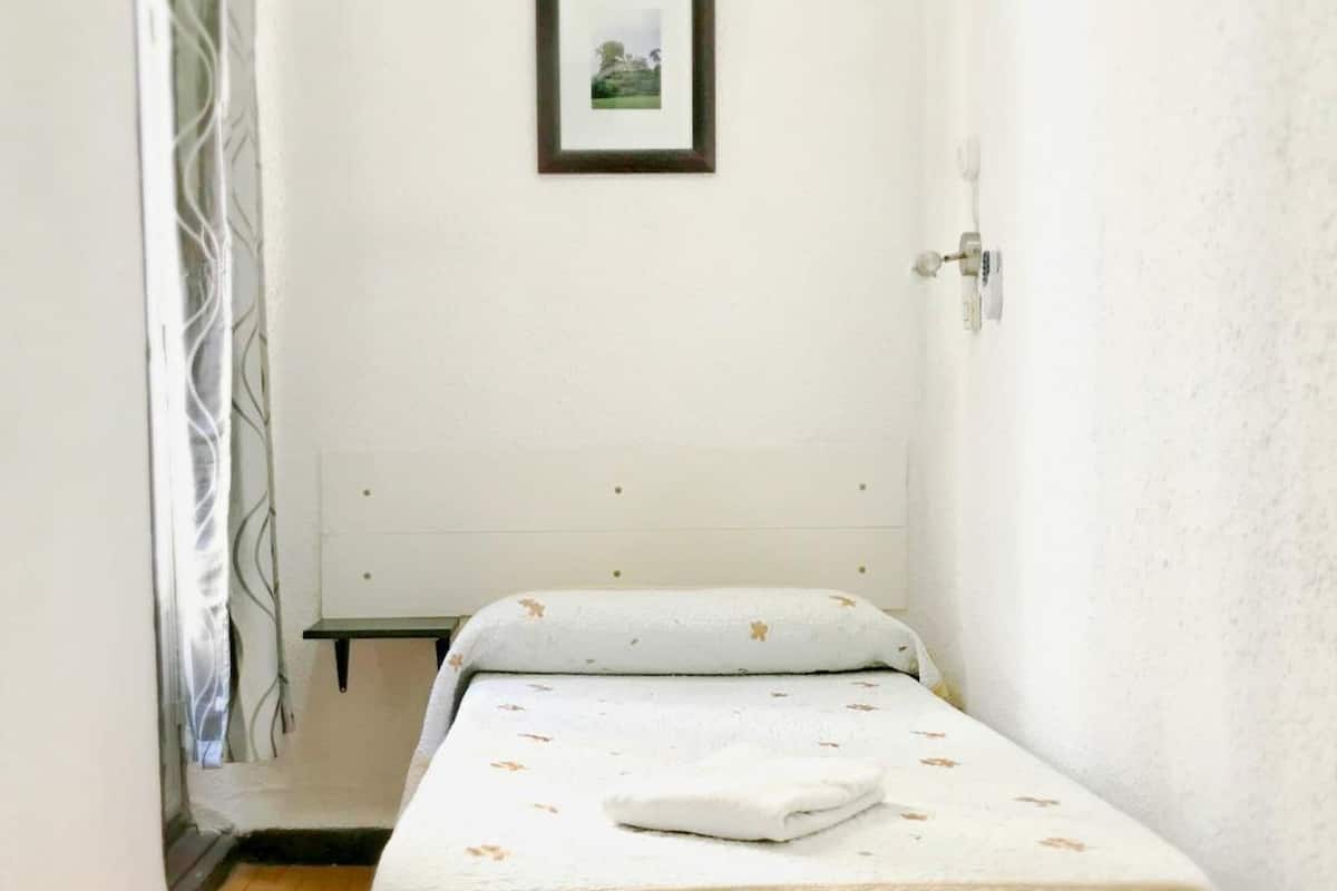 Um quarto pequeno e estreito, com uma cama de solteiro, um quadro na parede e uma porta com puxador.