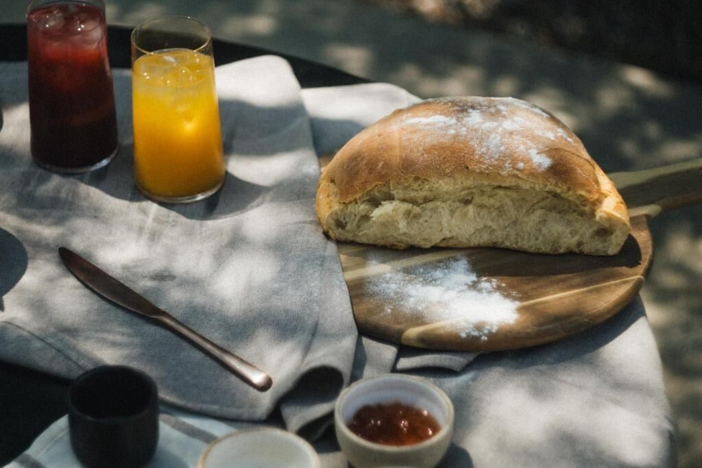 Uma cesta de piquenique com pão numa tábua de madeira, uma faca, um copo de suco de laranja e um copo de bebida vermelha sobre um tecido.