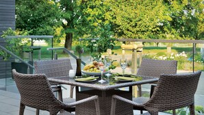 Mesa de vime posta com uma travessa de frutas, copos de vinho e talheres numa varanda com vista para vegetação exuberante.