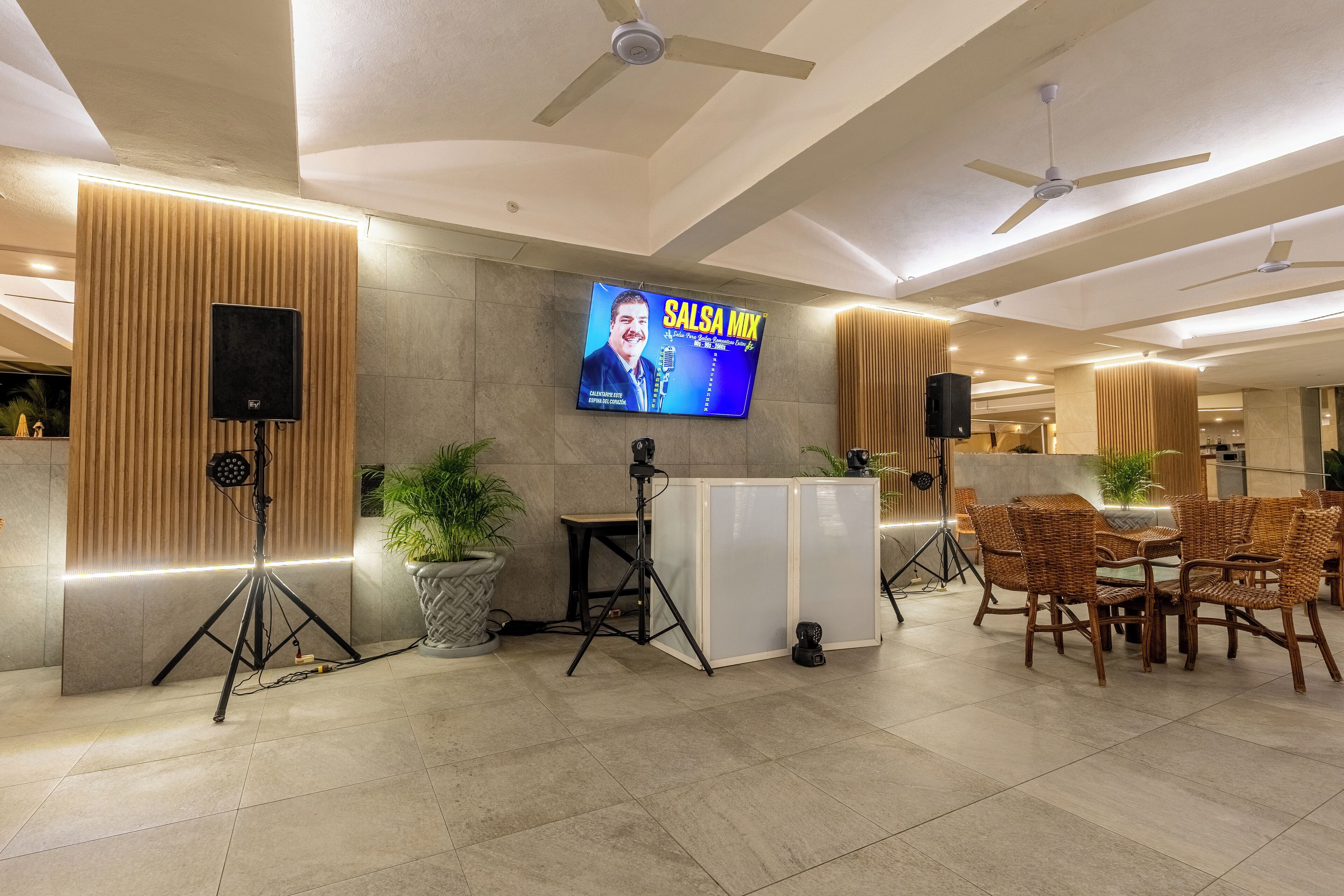 Um lobby moderno com uma TV exibindo 'Salsa Mix', uma mesa de recepção, cadeiras de vime e ventiladores de teto.