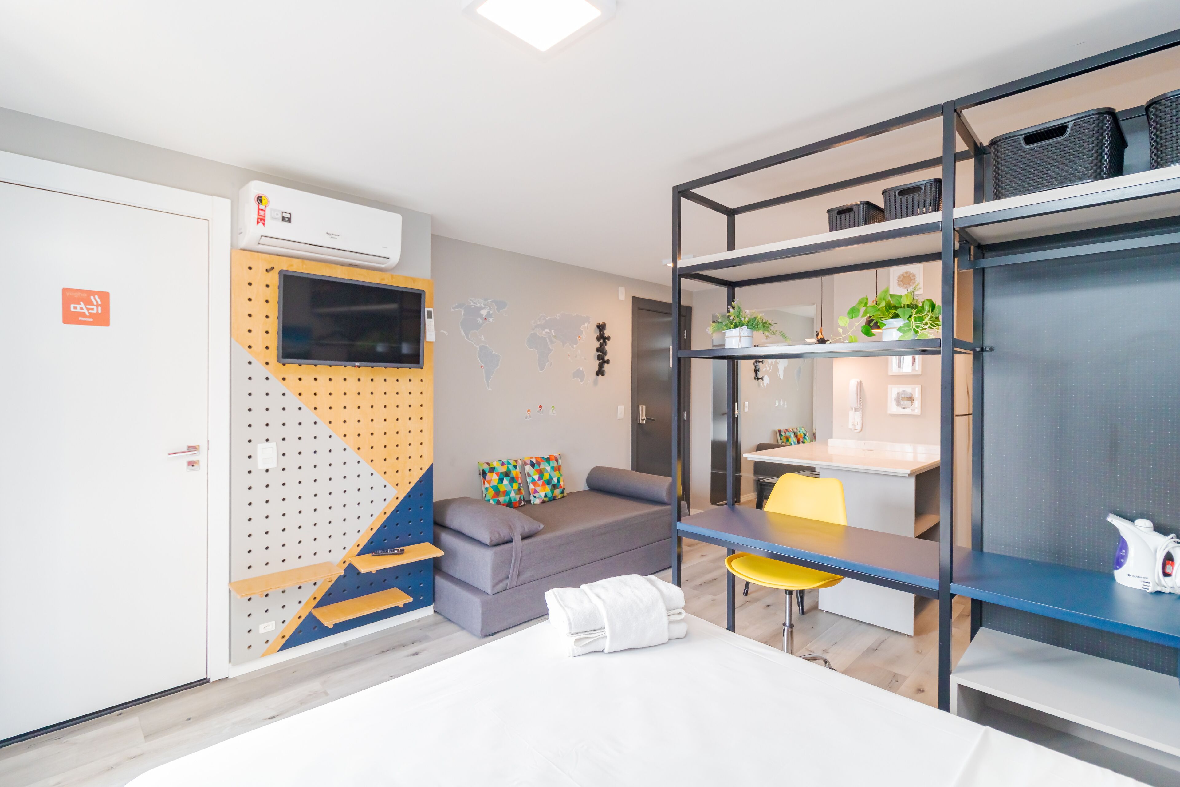 Quarto de hotel moderno com um sofá cinzento, uma pequena mesa com uma cadeira amarela, uma TV montada na parede e um mapa-múndi decorativo.
